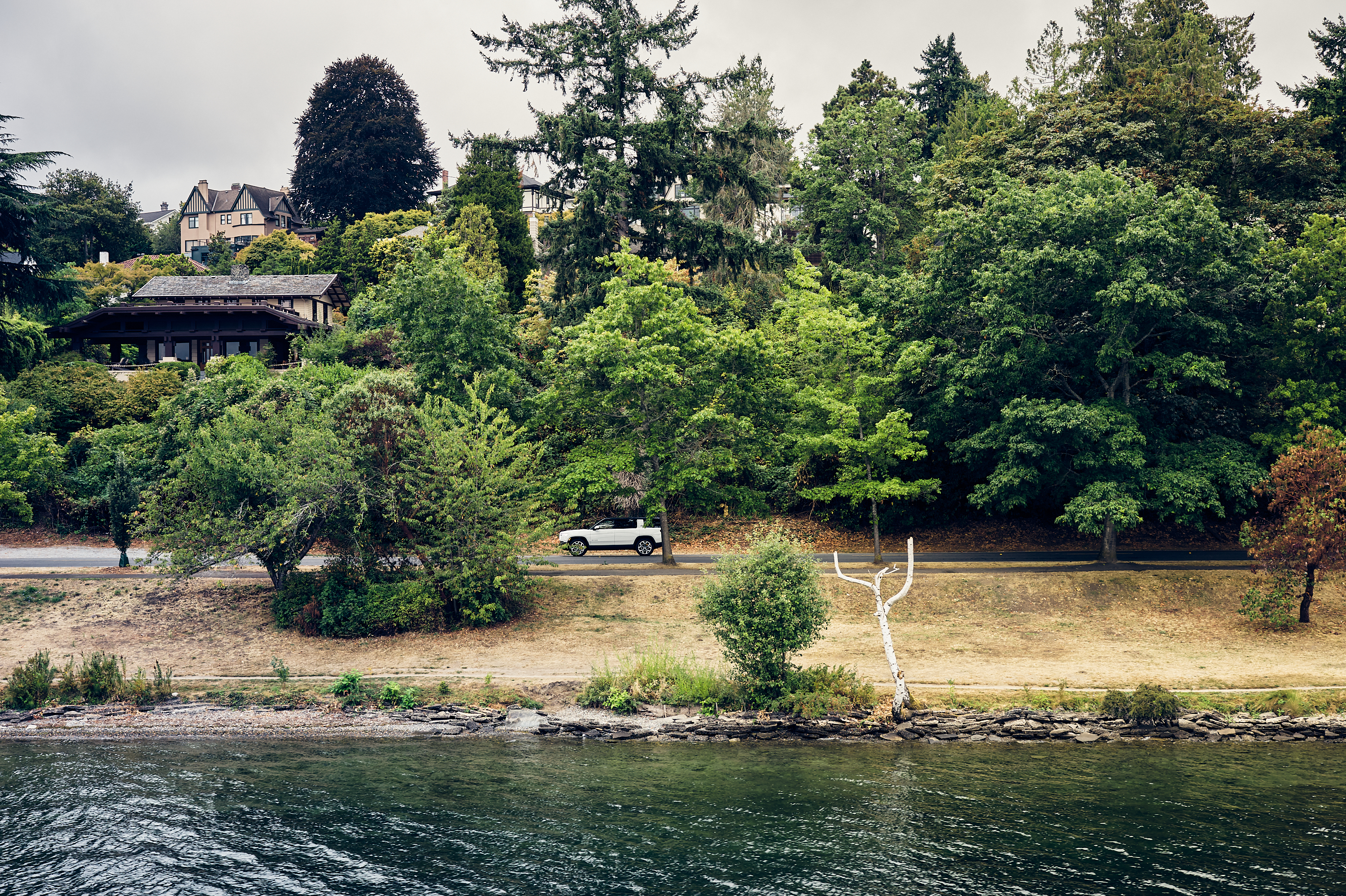 White Rivian R1 drives along a road lined with trees and houses, viewed ...