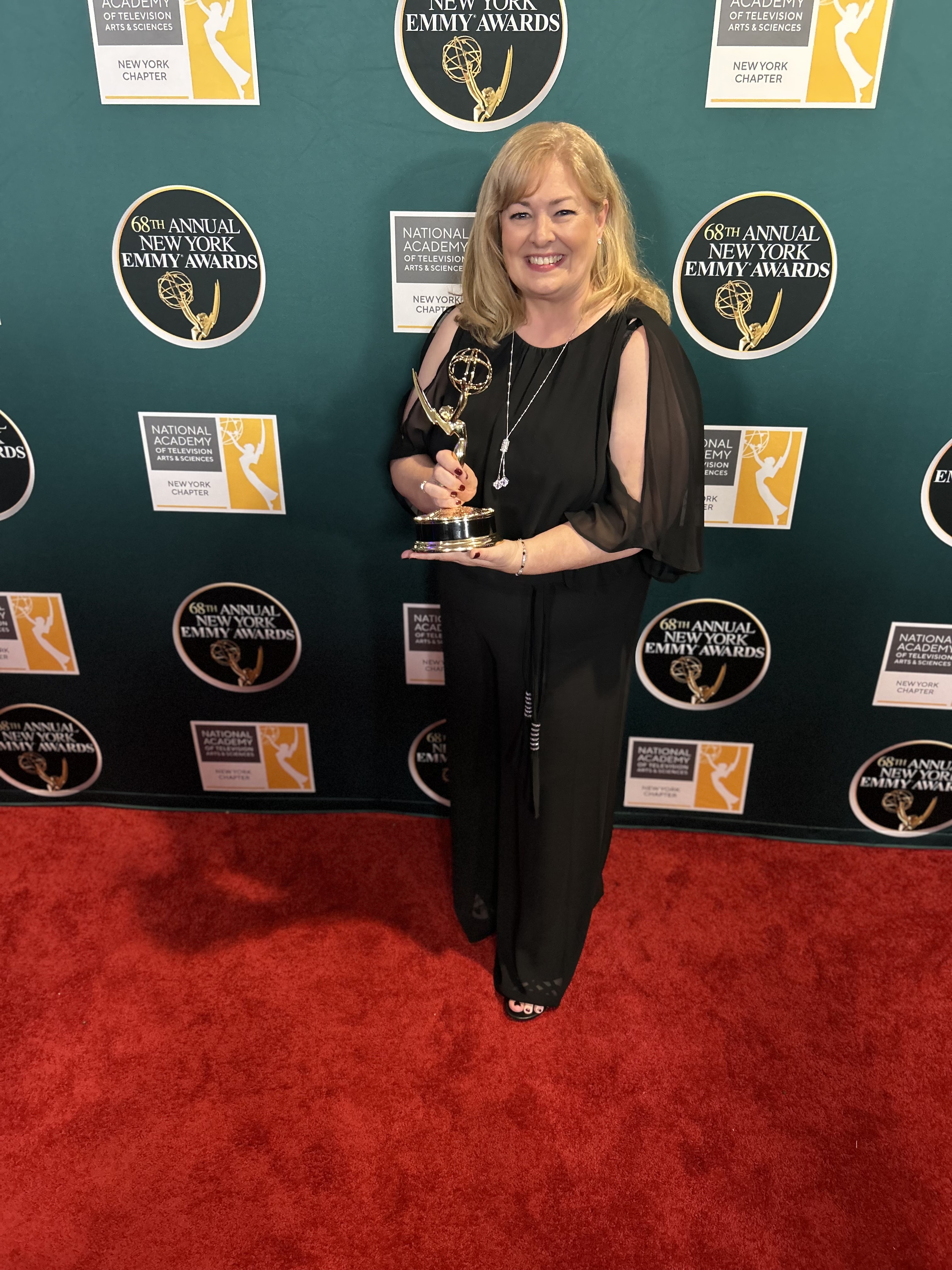 Woman smiles, holding Emmy award on red carpet in front of
