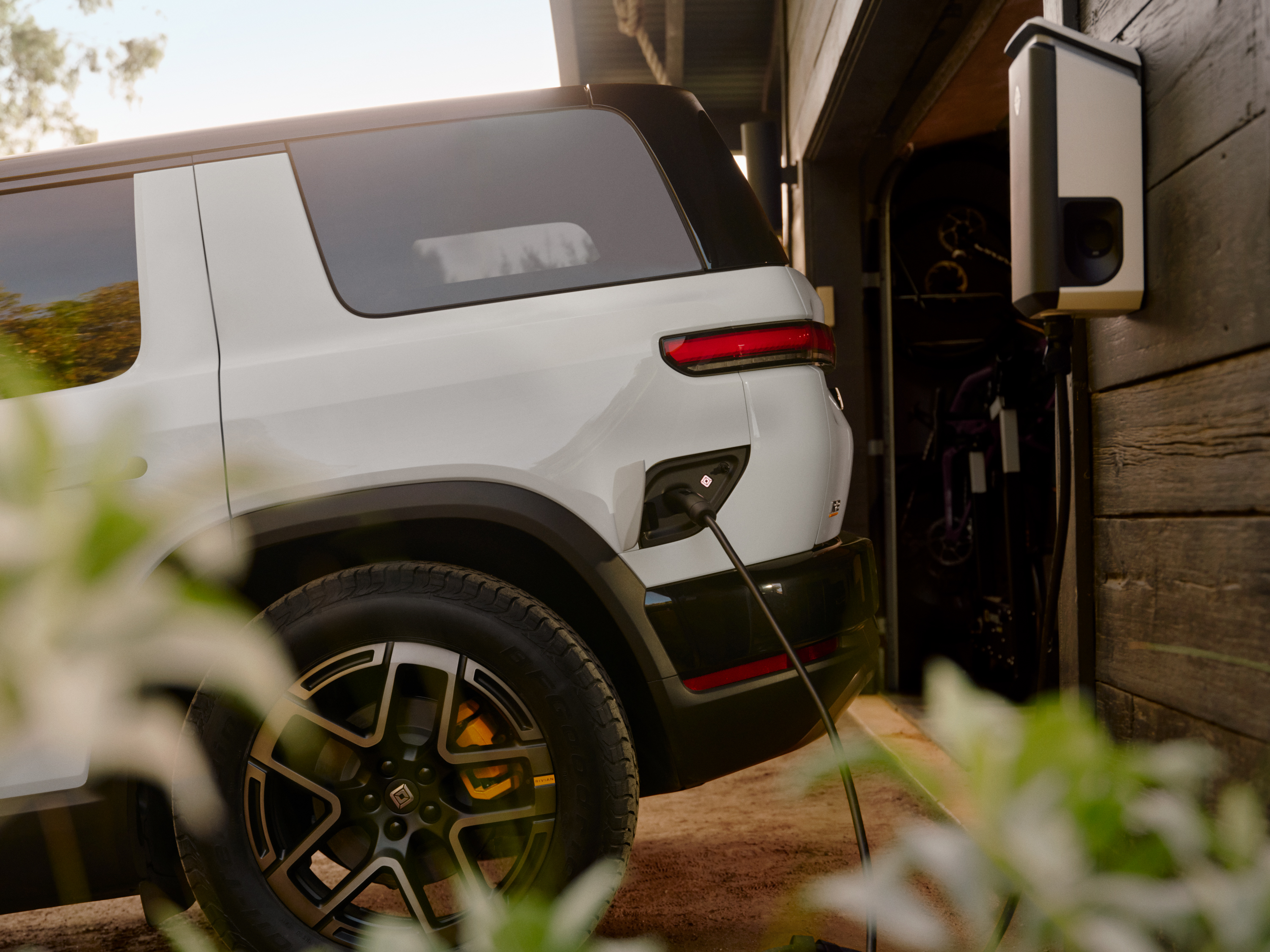 White Rivian SUV charges via a wall-mounted charger outside a garage. A ...