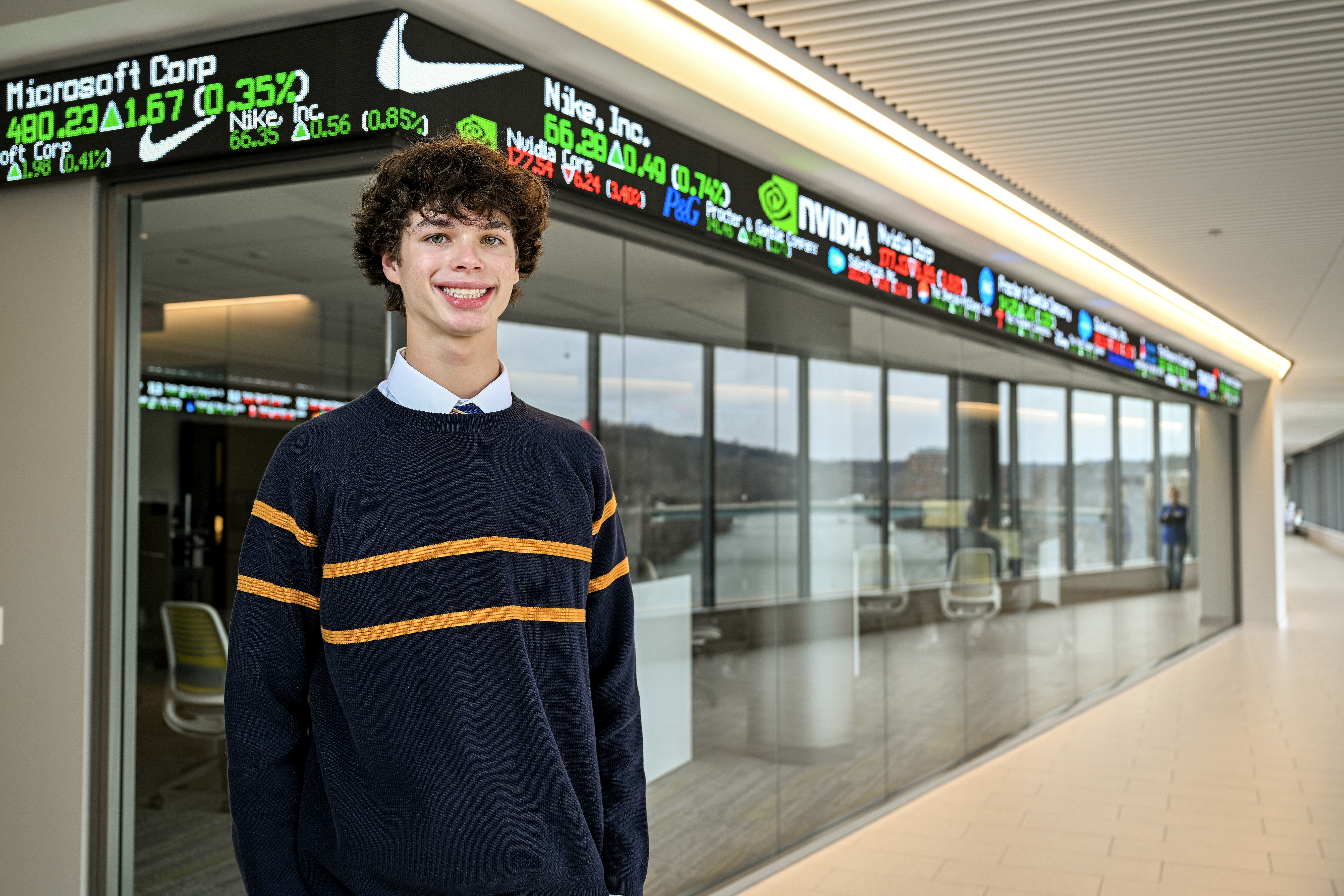 Teen smiles in front of stock ticker. He wears a navy sweater with gold ...