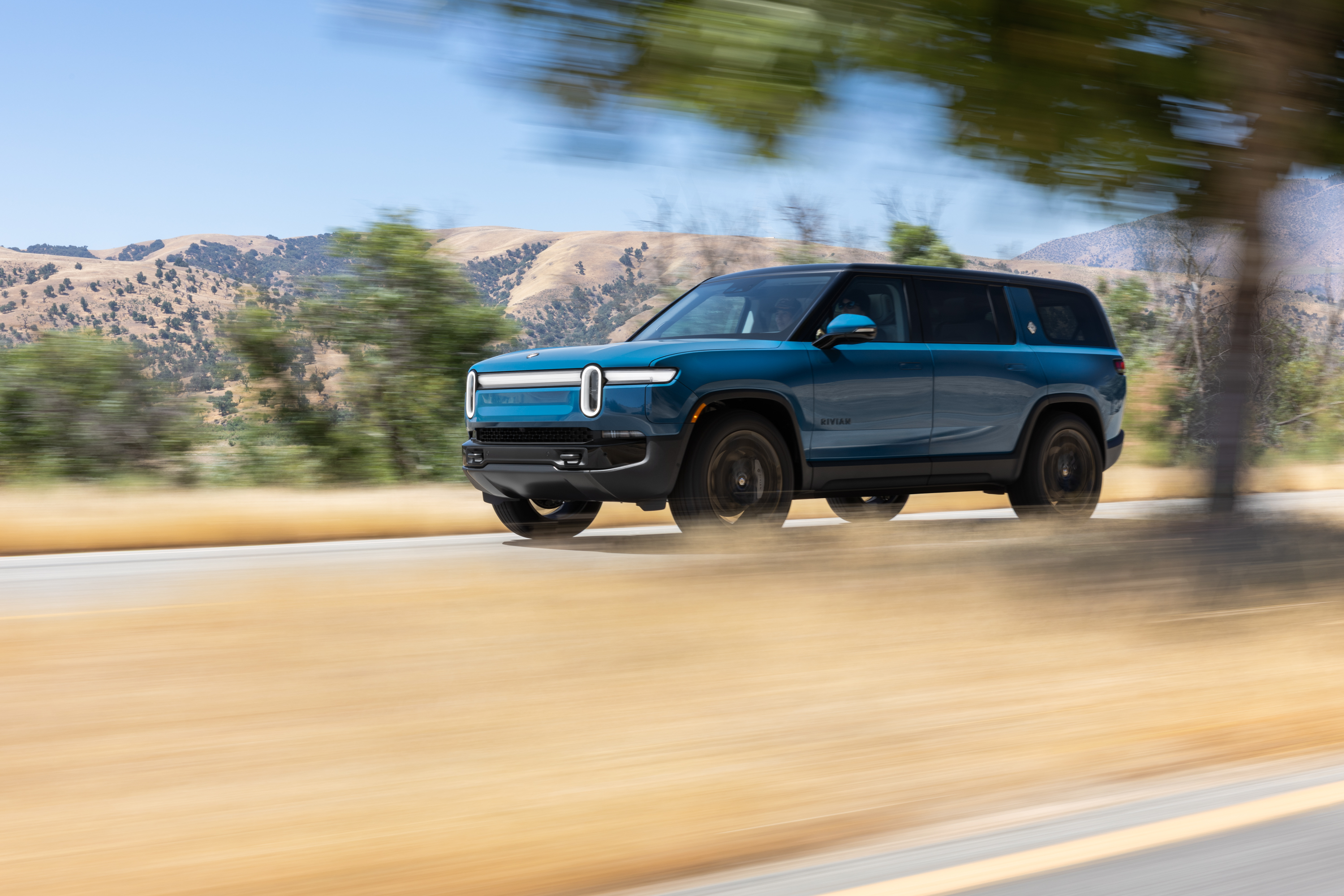 A blue Rivian SUV drives on a road. The background shows rolling hills ...