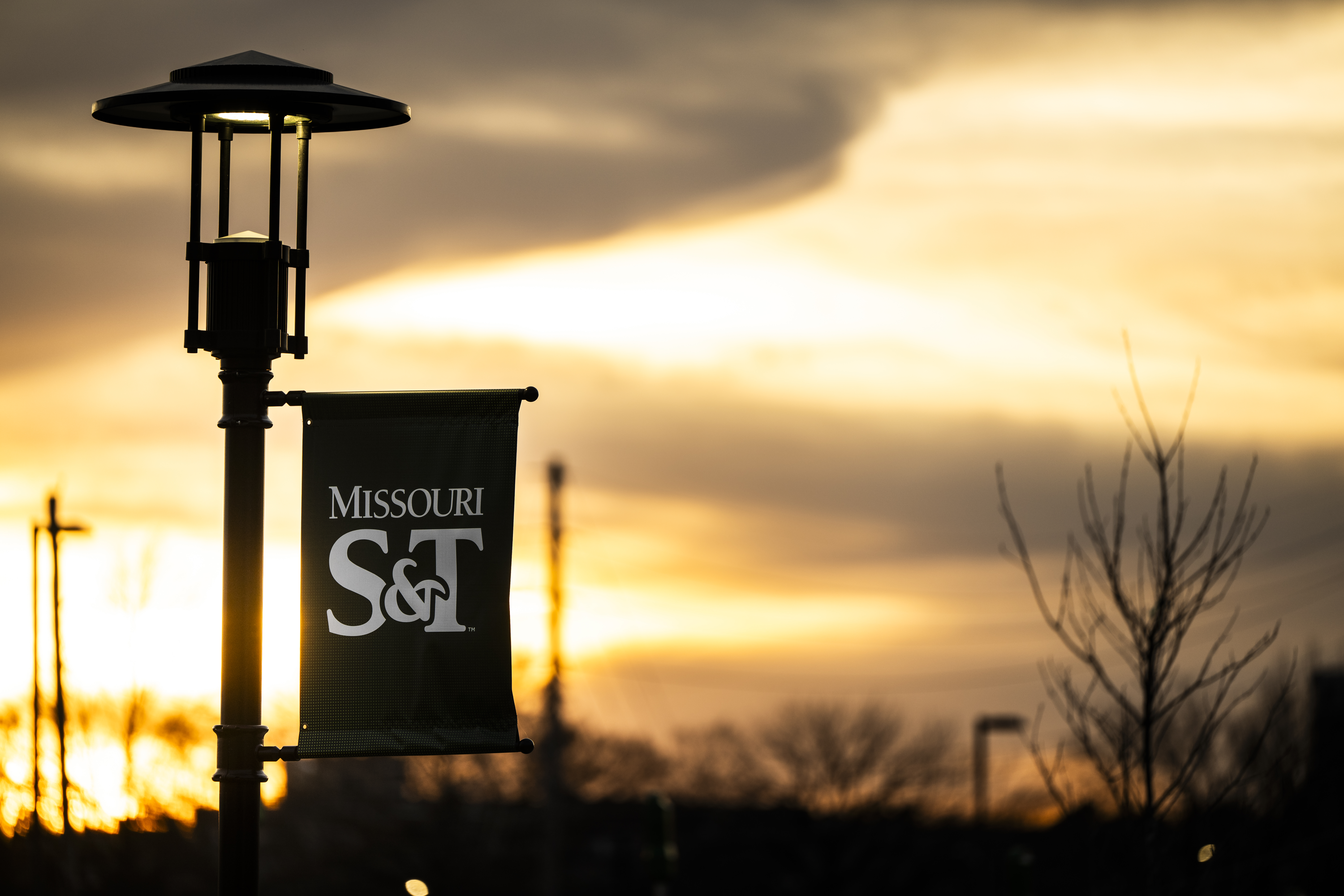 Streetlight displays a Missouri S&T banner against a sunset sky. The ...