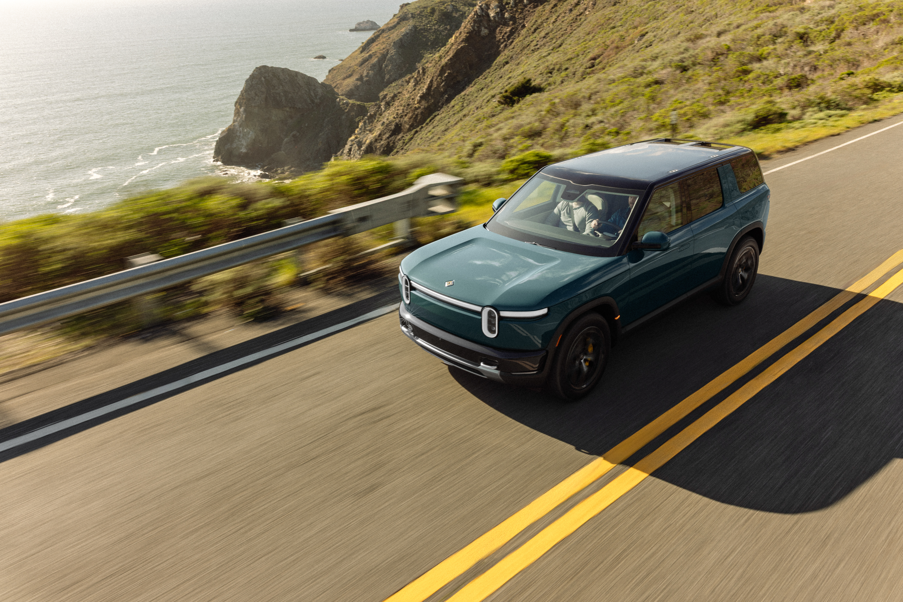 A teal Rivian SUV drives on a highway along a coastline. The vehicle ...