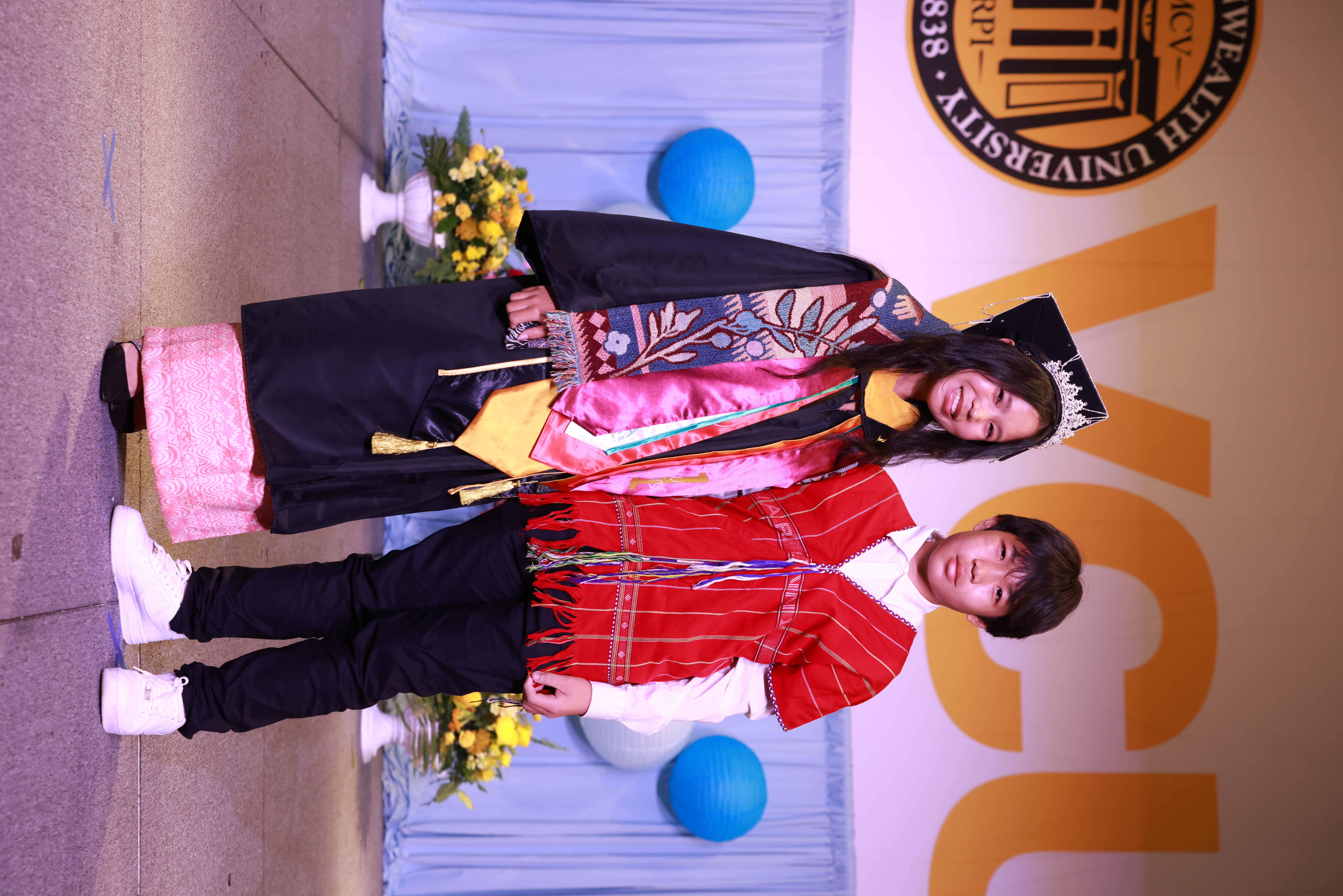 Graduate in cap and gown poses with a person in a red shirt on a stage ...
