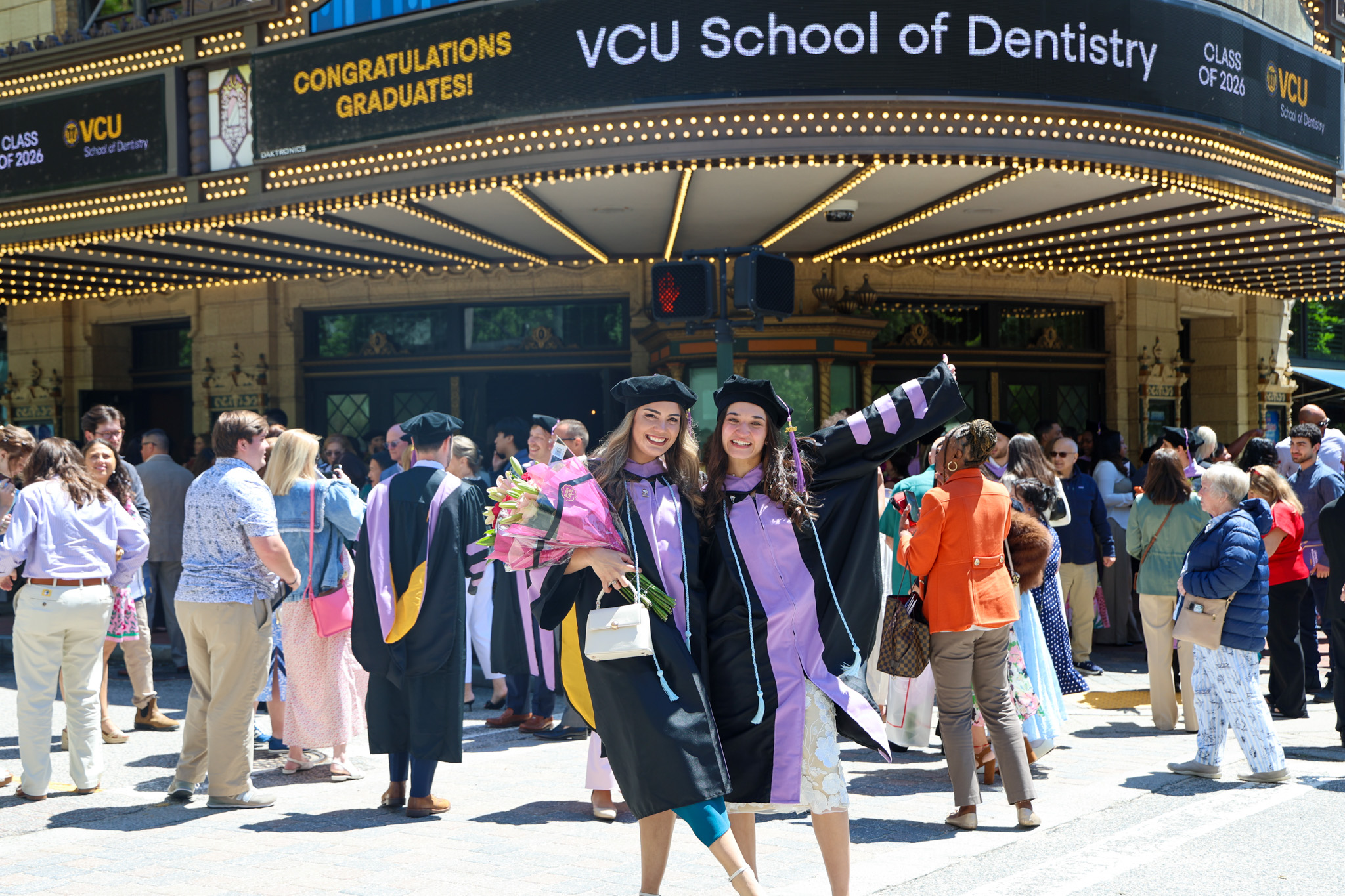 Two graduates pose for a photo in front of the theater with a crowd of ...