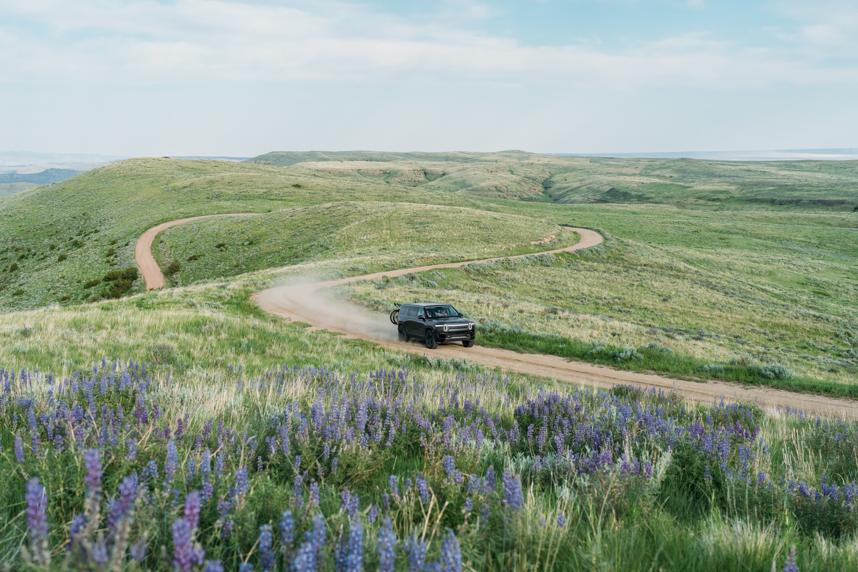 Rivian SUV drives on a dirt road through a grassy, hilly landscape. The ...