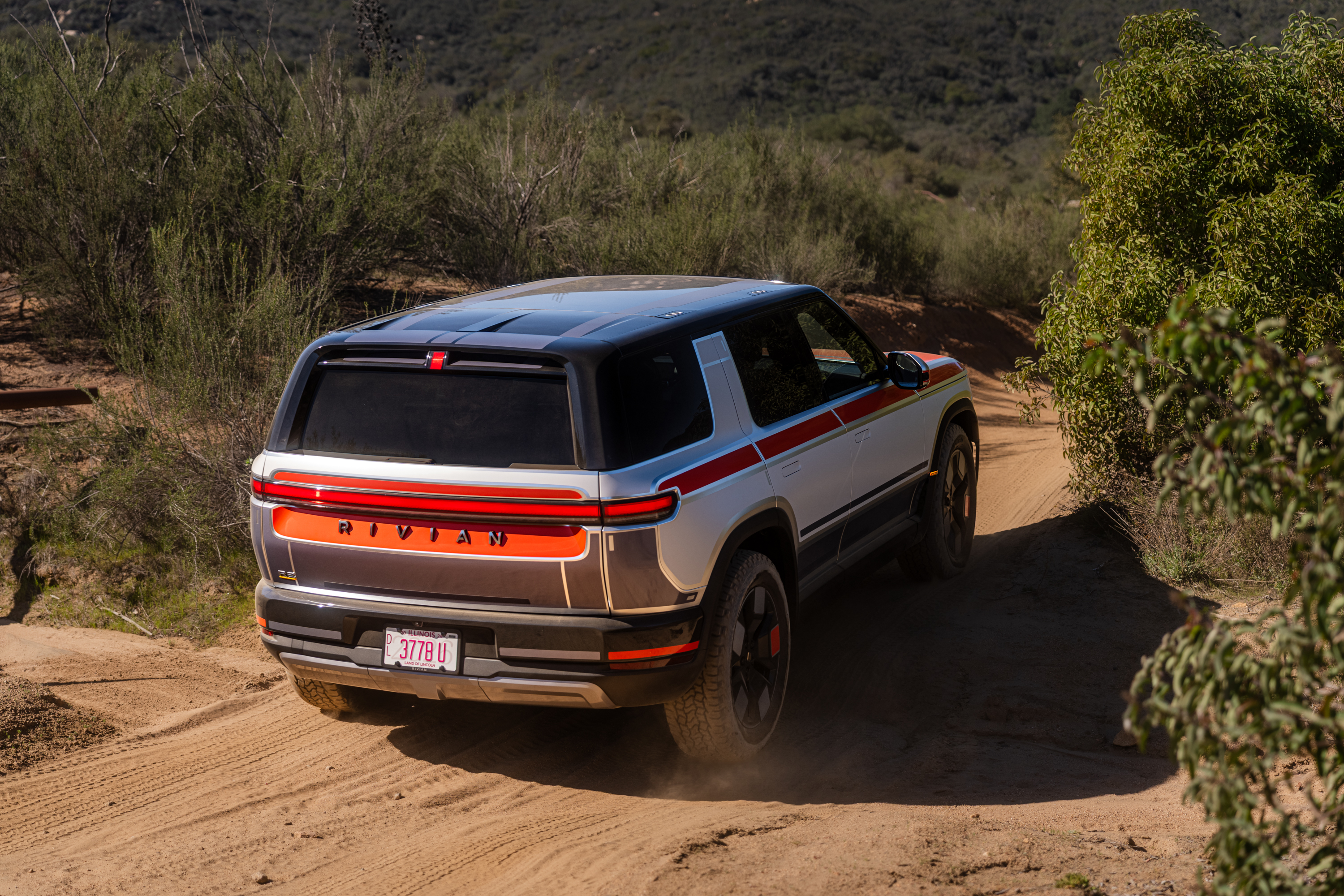 A Rivian SUV drives on a dirt road. The vehicle is white with a red ...
