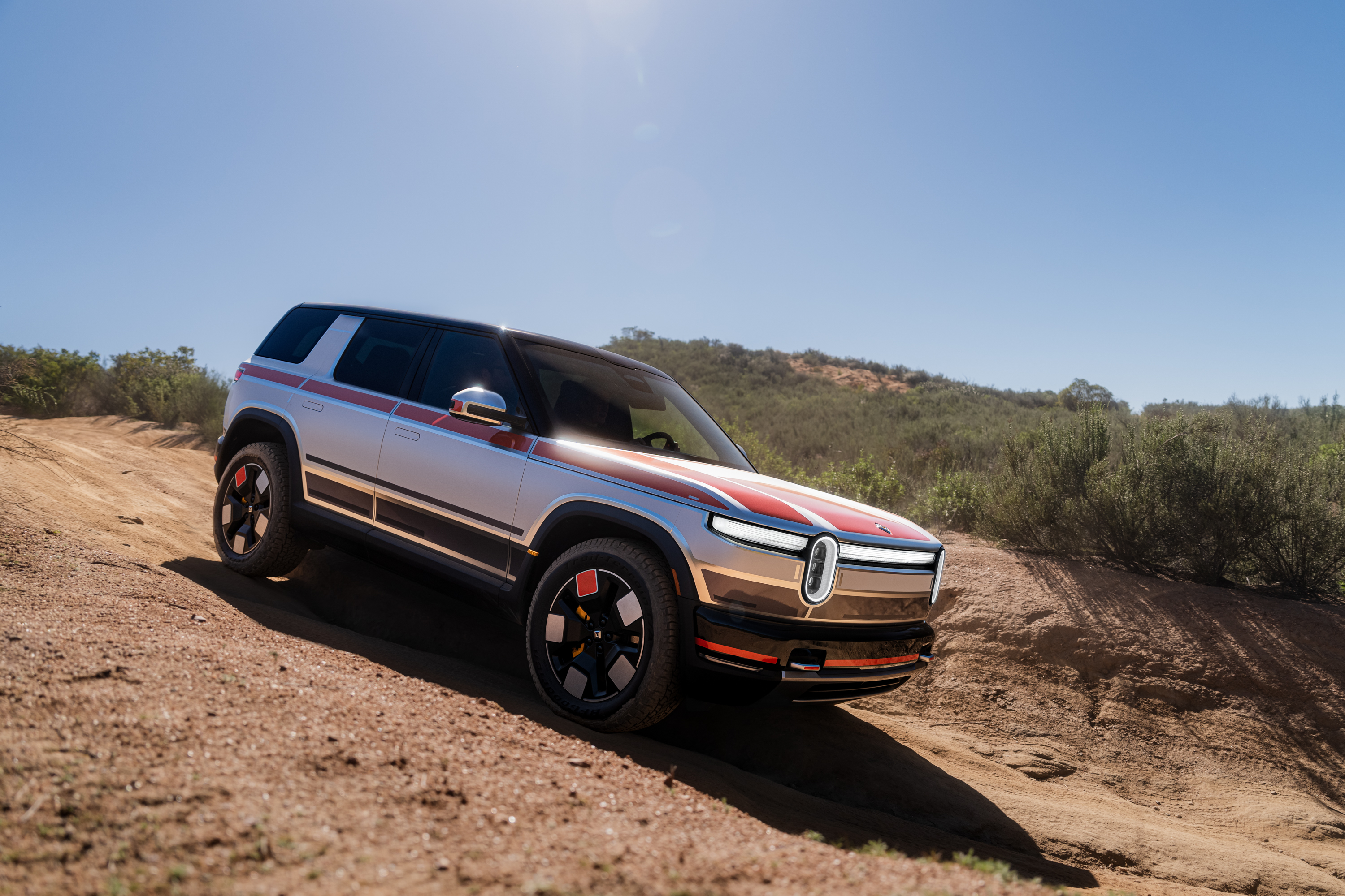 Rivian SUV drives on a dirt road. The vehicle is silver with red and ...