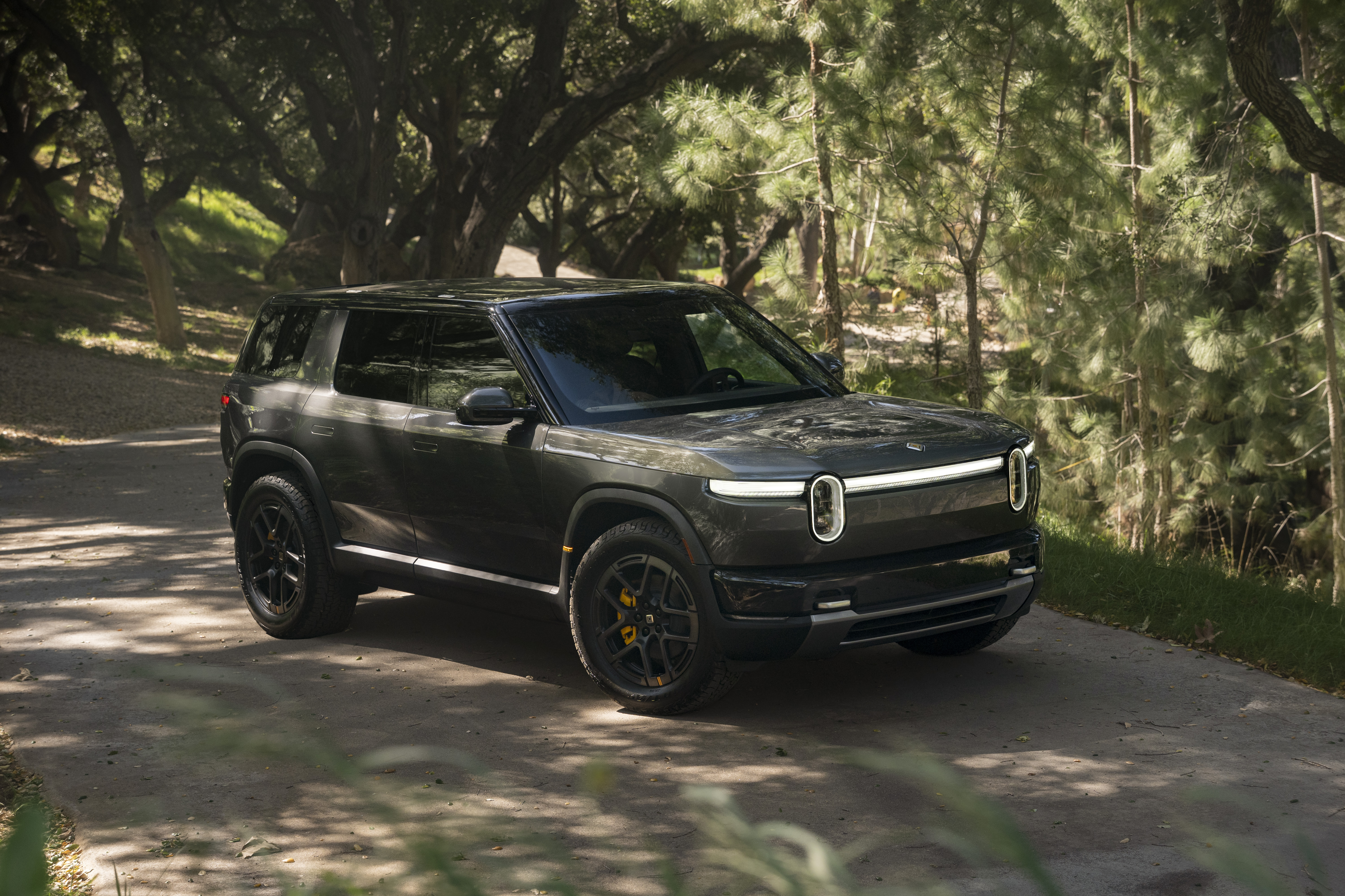Rivian R2, a dark SUV, parks on a tree-lined road. The vehicle has a ...